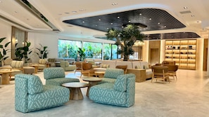 Espaço de convívio moderno com sofás, cadeiras e uma mesa de centro, com uma grande janela e vista para o verde.