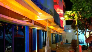 Exterior de hotel à noite, com letreiros iluminados, galeria coberta e vasos de plantas.