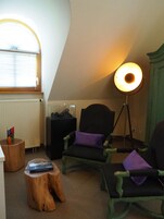 Um quarto com duas poltronas verdes, um banquinho de madeira, um abajur, um radiador e uma janela com persianas.