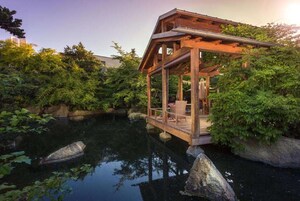 Um gazebo de madeira com estrutura de vidro, posicionado sobre um lago rodeado por vegetação exuberante.