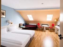 Quarto de hotel com cama grande, escrivaninha, cadeira e mesa pequena. Duas janelas oferecem luz natural.