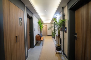 Um corredor com portas de madeira, um banco, plantas em vasos e um mostrador digital de elevador.