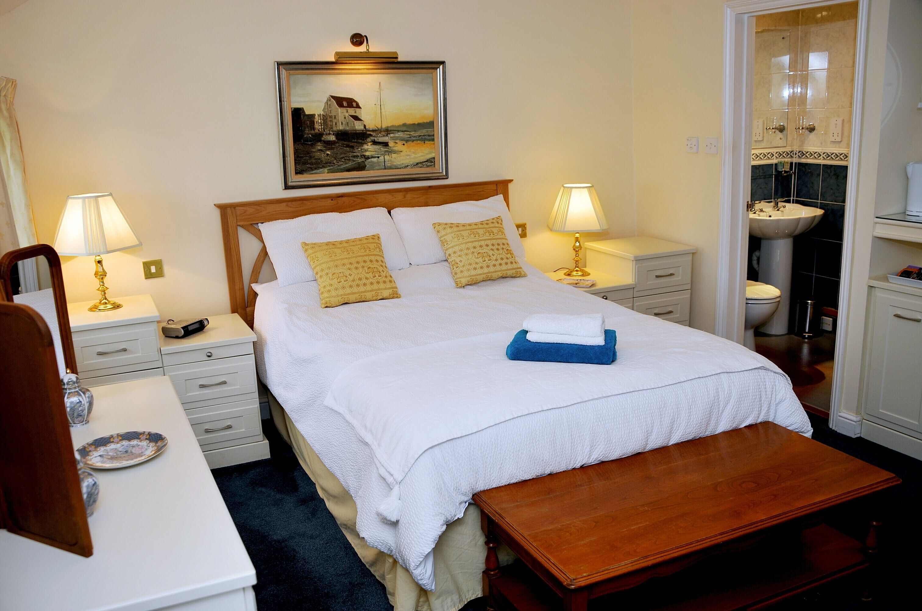 Quarto de hotel bem arrumado, com cama, mesas de cabeceira, escrivaninha e banheiro visível por uma porta aberta.