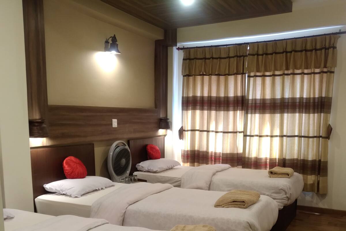 Quarto de hotel com quatro camas, cada uma com colcha branca e travesseiro vermelho. Há um aquecedor fixado na parede e um ventilador. O quarto tem piso de madeira e uma janela com cortinas listradas.
