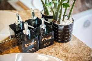 Uma bancada de banheiro com dosadores de sabonete pretos identificados como "One Hotel" e um vaso de planta com pedrinhas brancas.