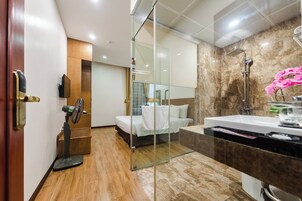 Casa de banho moderna com cabine de chuveiro em vidro, uma sanita branca e uma cama visível através do vidro.