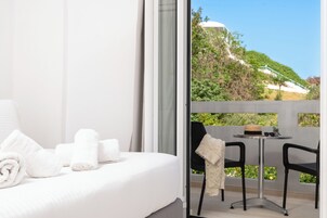 Quarto de hotel com cama, toalhas brancas, uma mesinha com chapéu e óculos de sol, e vista para uma encosta verde.