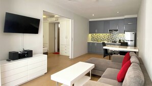Uma sala de estar moderna com uma televisão de tela plana, uma mesa de centro branca e um sofá cinza com almofadas vermelhas. Uma cozinha com armários cinza e painel de azulejos é visível ao fundo.