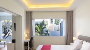 Quarto de hotel com cama, escrivaninha e cadeira. Há uma janela com vista para palmeiras e edifícios.