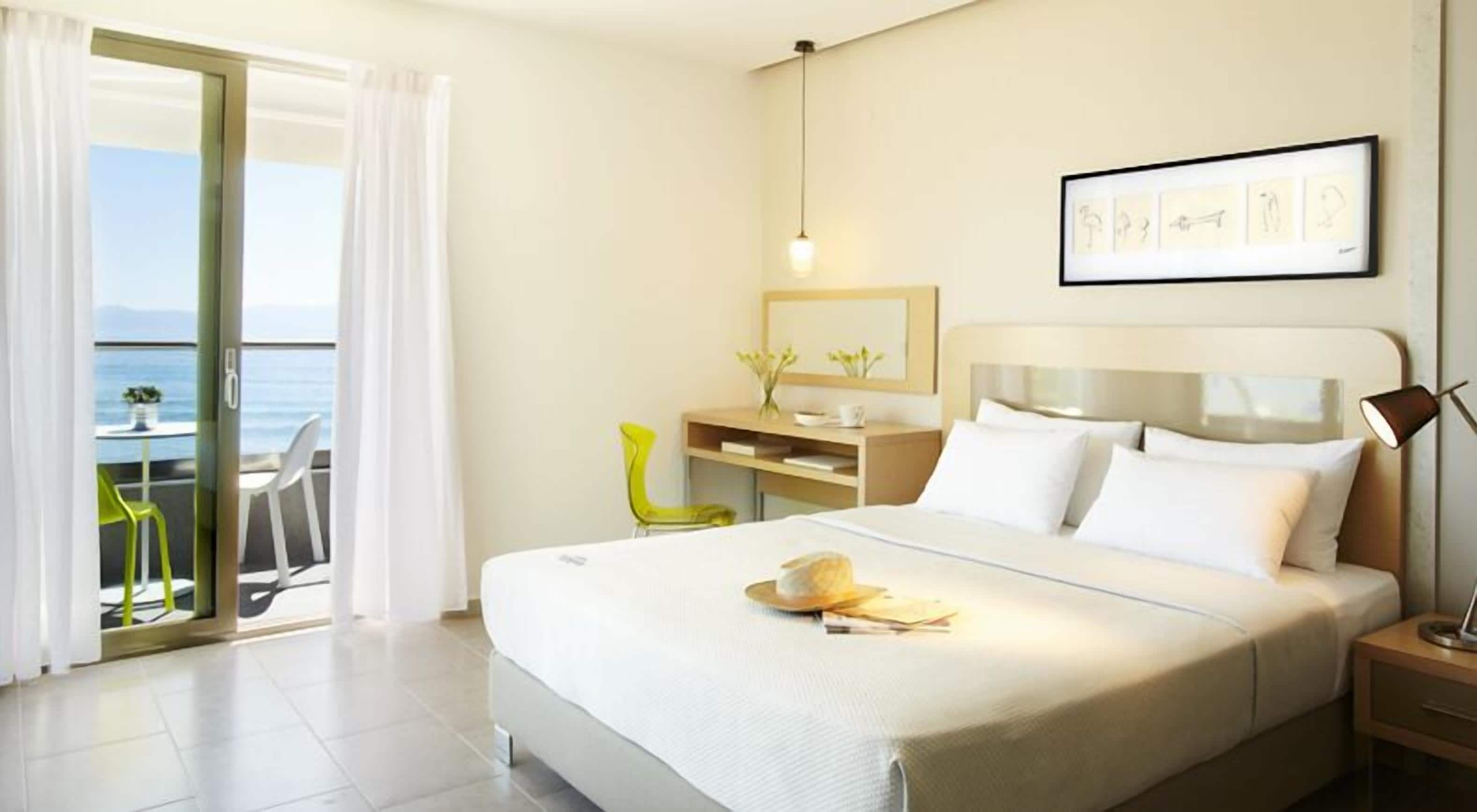 Quarto de hotel com uma cama, uma escrivaninha, uma cadeira, uma mesa de cabeceira, uma luminária, um quadro e vista para o mar através de uma porta de correr.
