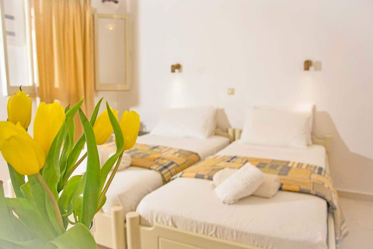Quarto de hotel com duas camas, um vaso de tulipas amarelas e uma janela com cortinas.