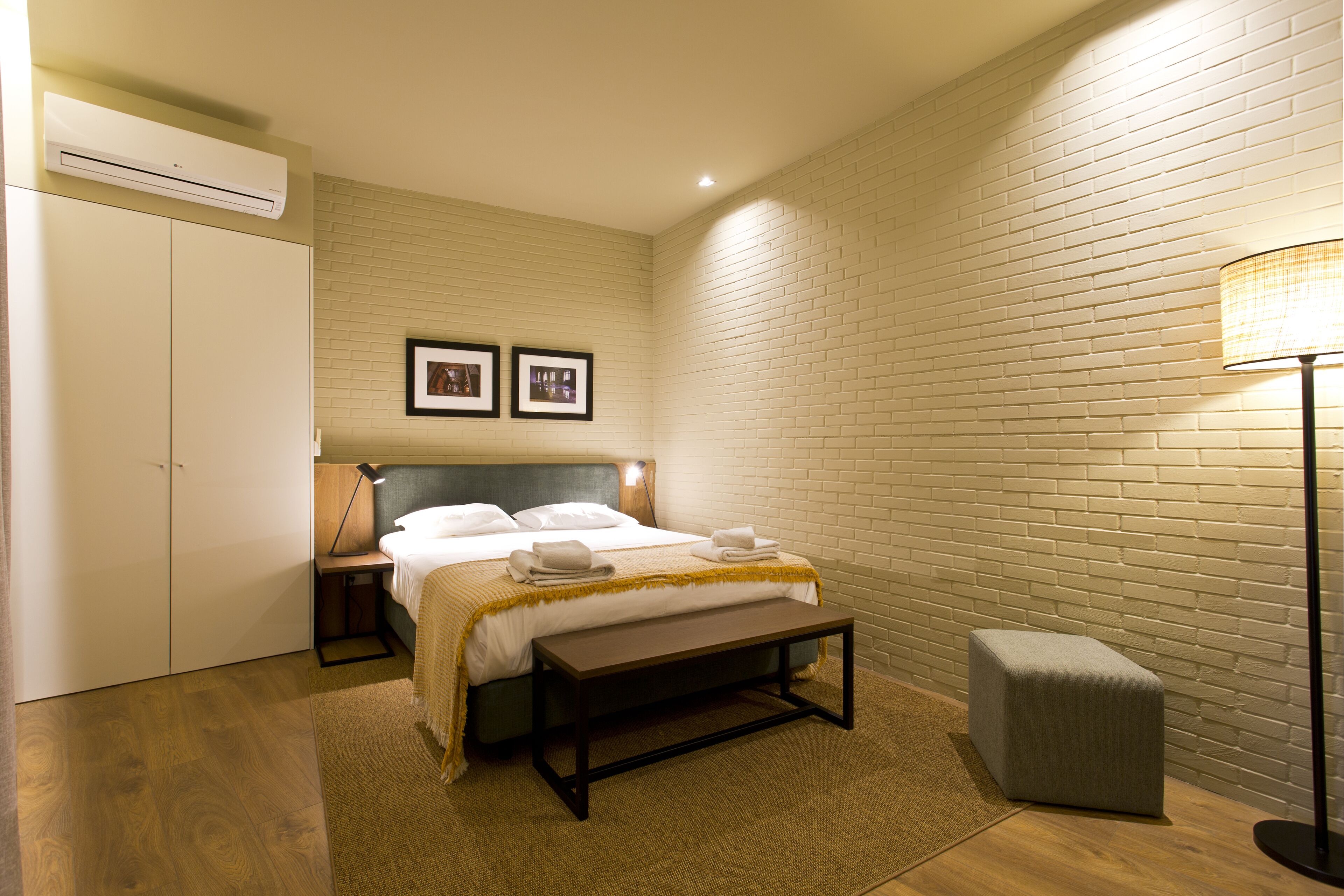Quarto de hotel com cama, mesa de cabeceira, abajur, banco e um pequeno banquinho.