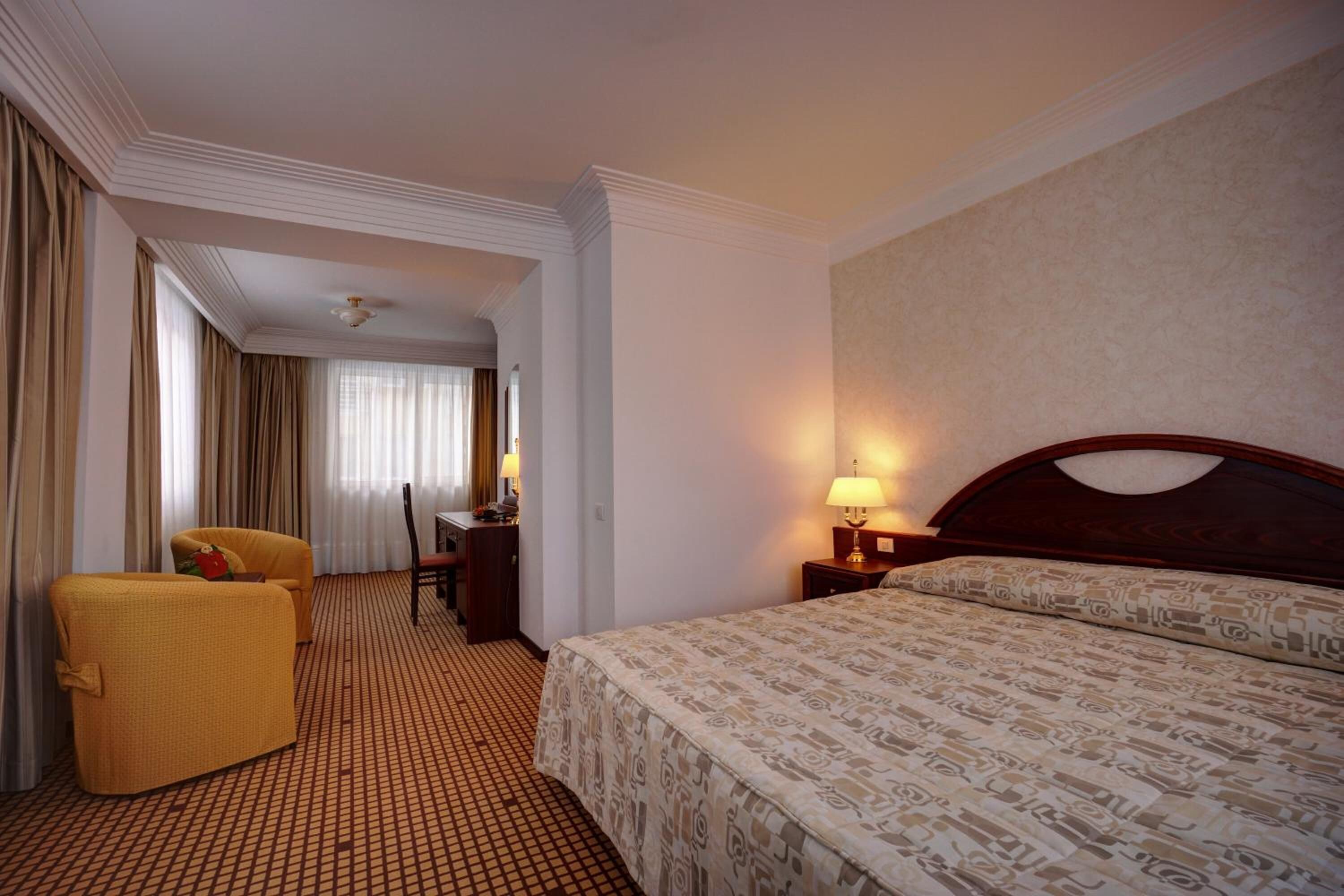 Quarto de hotel com uma cama grande, uma mesa com cadeira, um sofá pequeno e um tapete estampado.