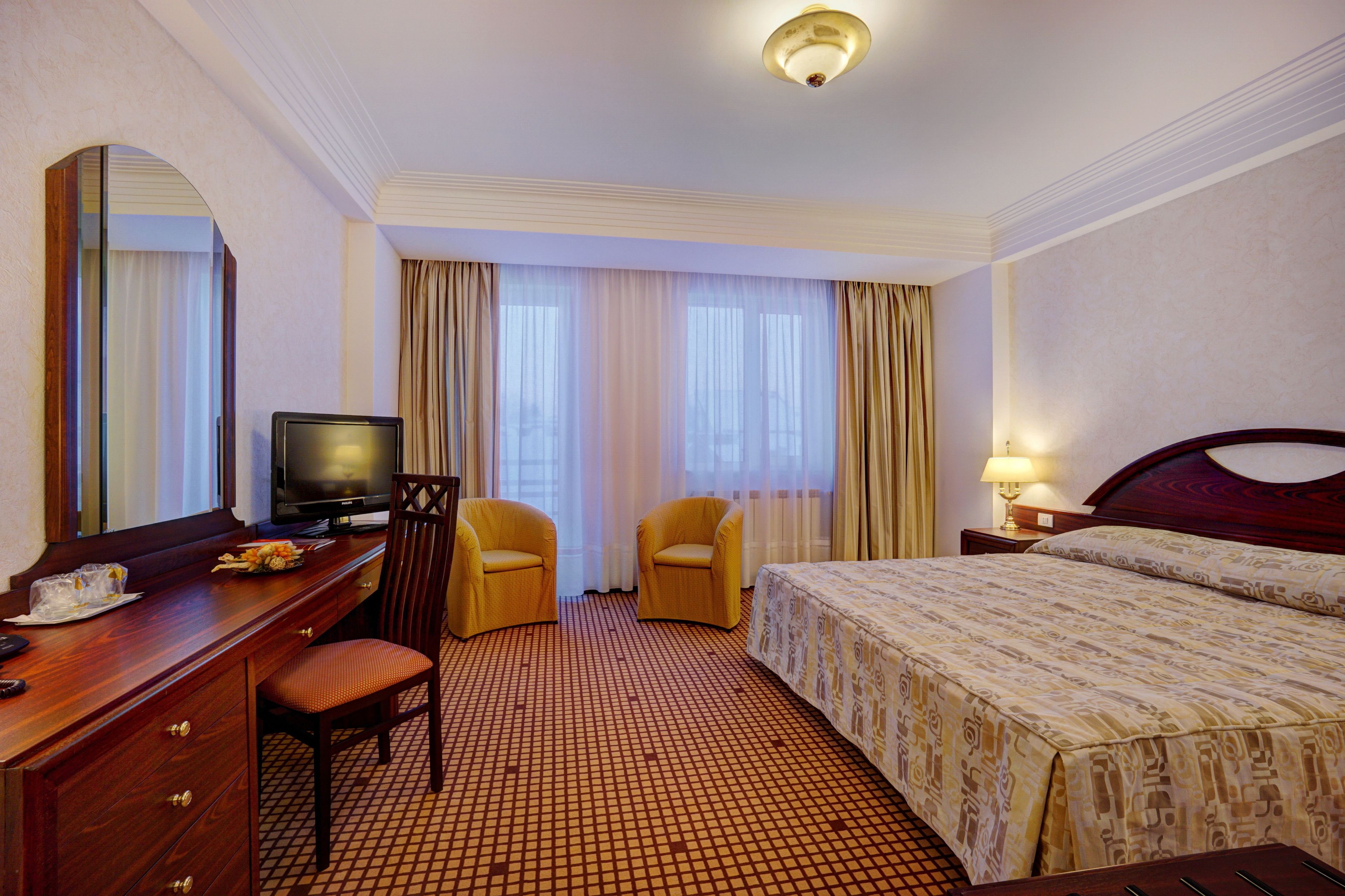Quarto de hotel com cama, uma escrivaninha com televisão, duas cadeiras e uma janela grande com cortinas.
