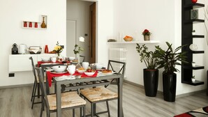Uma sala de jantar moderna com uma mesa de vidro, cadeiras pretas e toalha de mesa vermelha. Há plantas em vasos e uma estante preta com objetos decorativos.