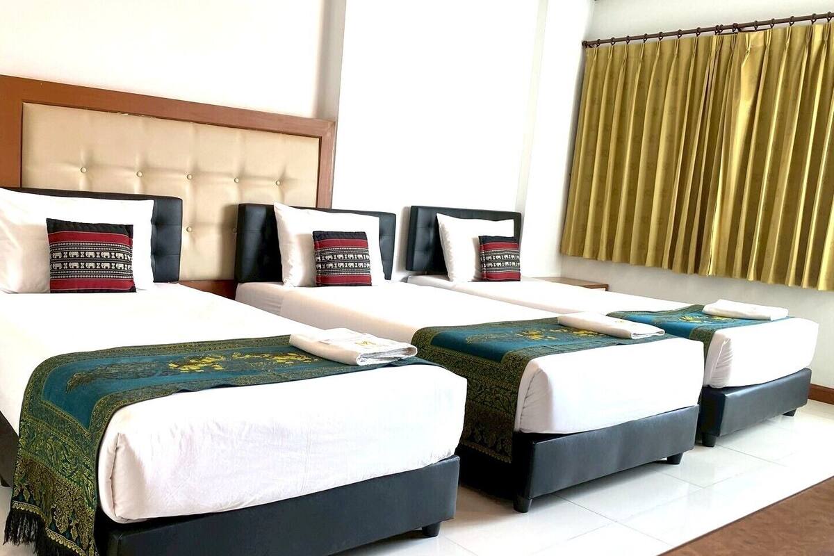 Quarto de hotel com três camas, cabeceira de madeira, mesa de cabeceira com abajur e janela com cortinas.