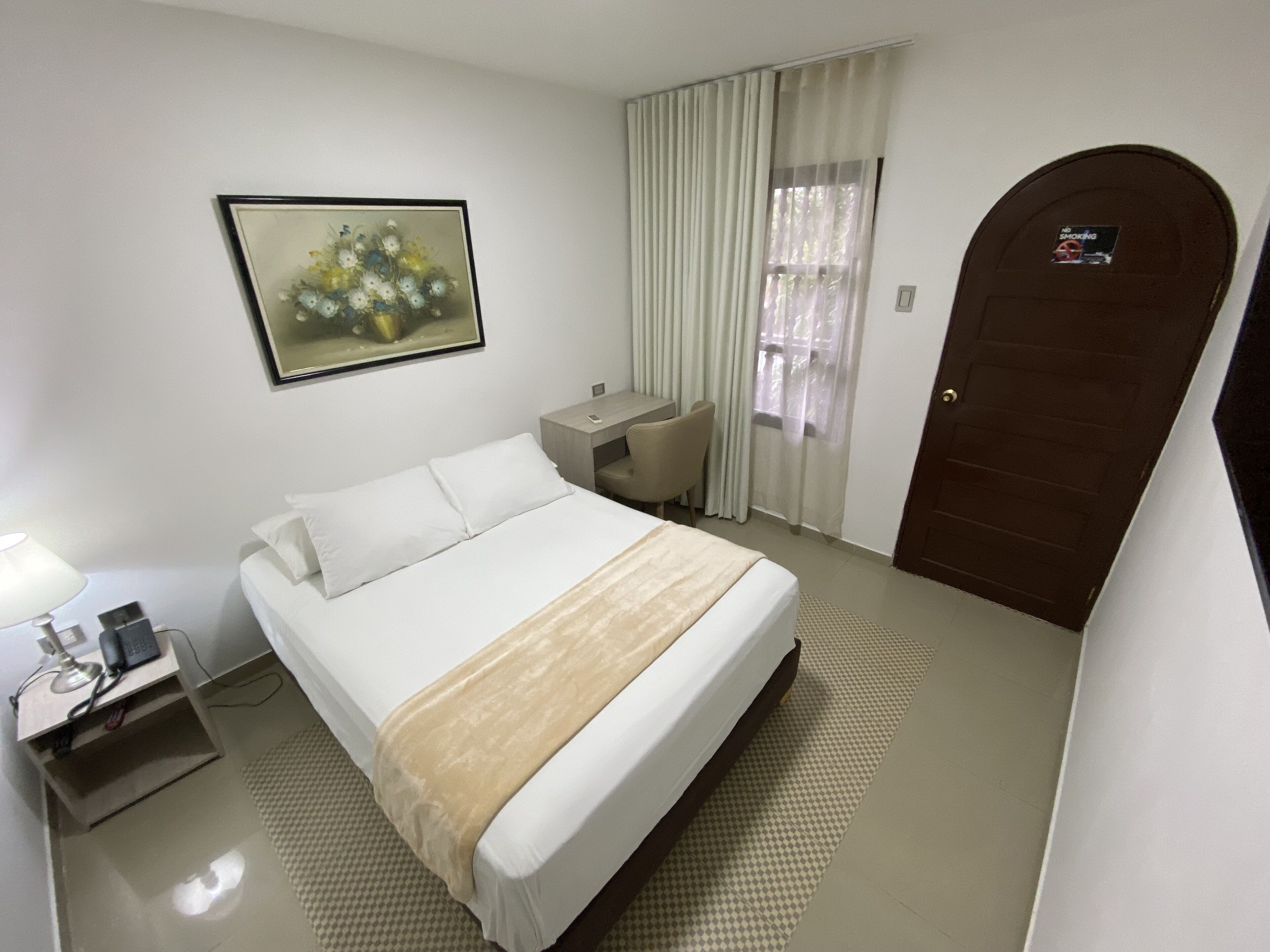 Quarto de hotel com cama, escrivaninha, cadeira e porta com placa de 'Não Fumar'.