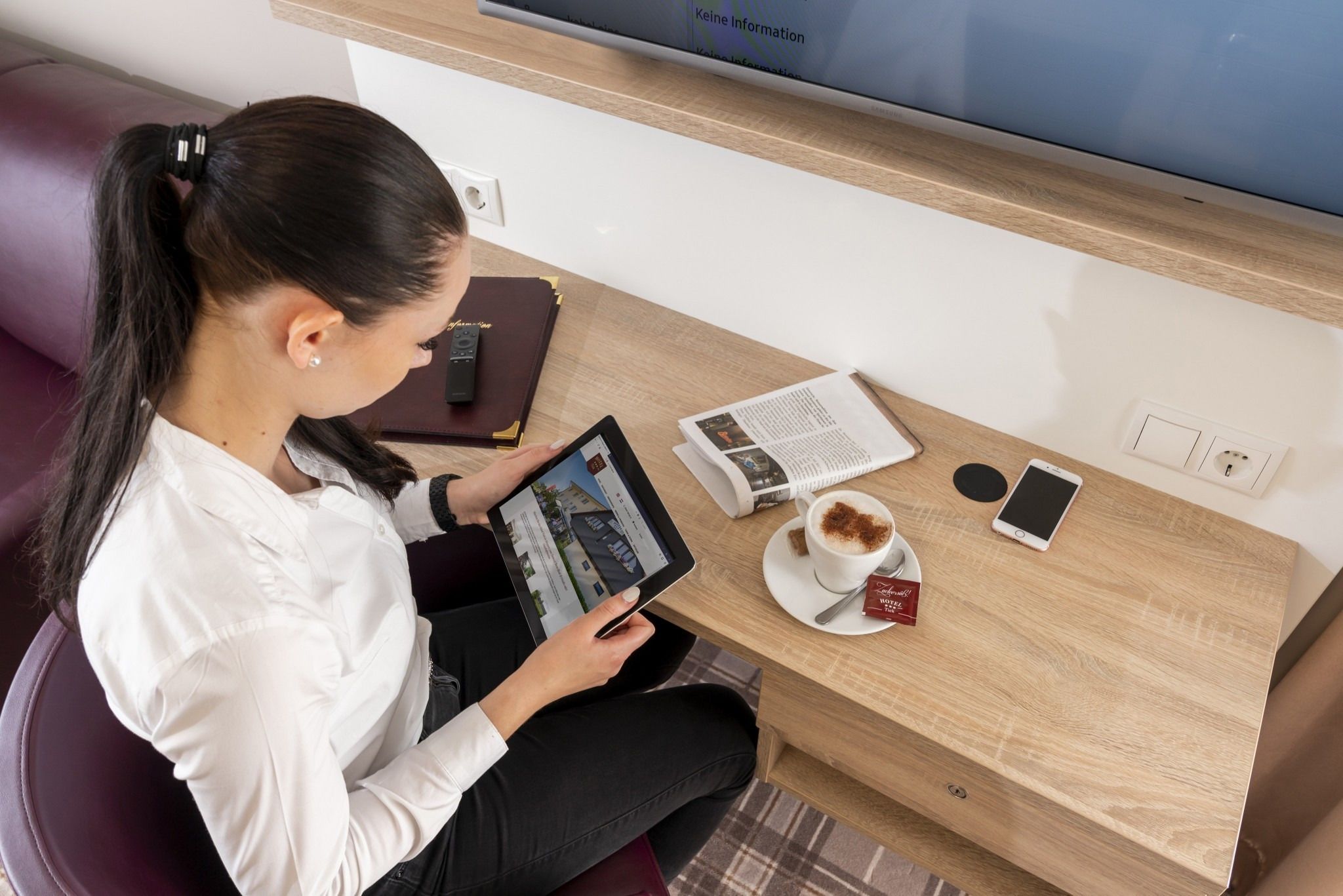 Uma pessoa sentada a uma mesa de madeira com um tablet, um smartphone, uma chávena de café e um bloco de notas.