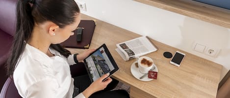 Uma pessoa sentada a uma mesa de madeira com um tablet, um smartphone, uma chávena de café e um bloco de notas.