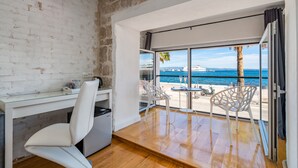 Quarto moderno com parede de tijolos brancos, escrivaninha de vidro, cadeira branca e varanda com vista para o mar e uma palmeira.