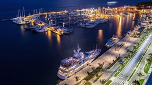 Marina com vários iates atracados, um iate grande em primeiro plano e um calçadão bem iluminado à beira-mar.