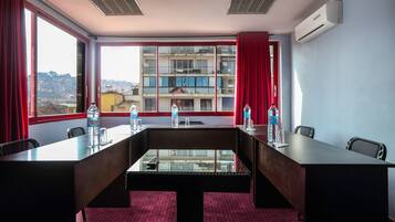 Sala de conferências com uma mesa comprida, cadeiras e vista para um edifício através de grandes janelas.