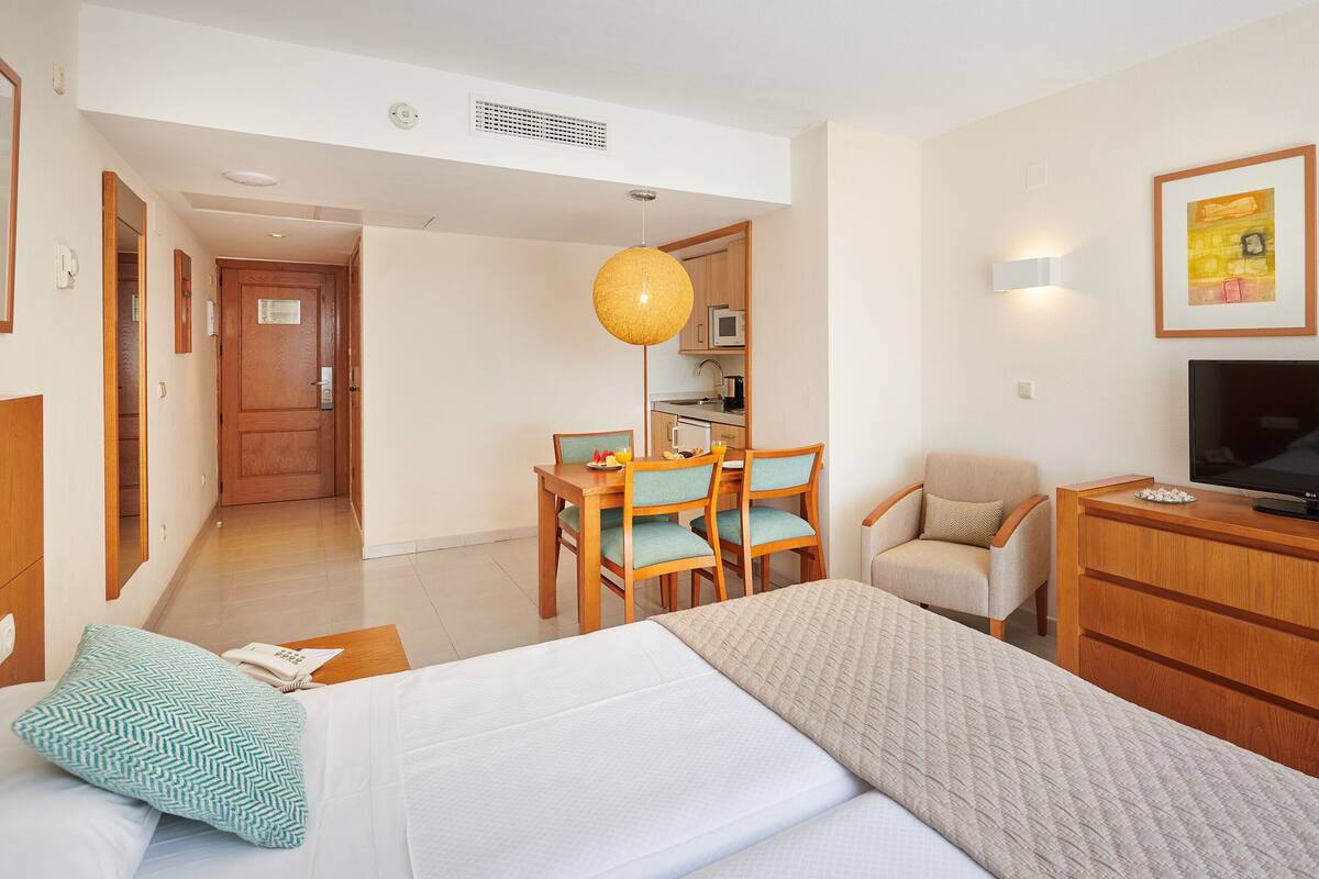 Quarto de hotel com cama, escrivaninha com cadeiras, televisão e cozinha compacta.