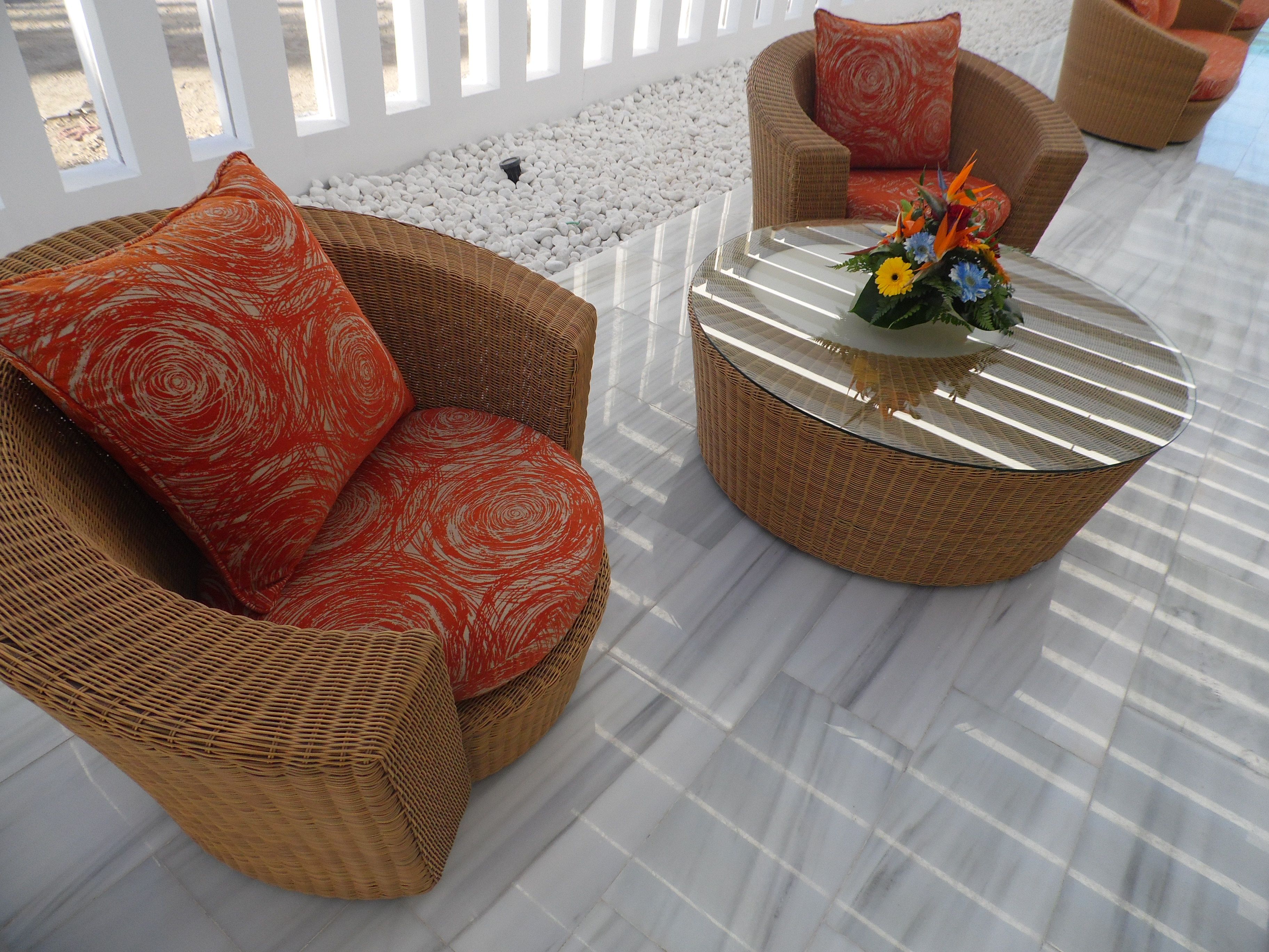 Um sofá de vime com almofada estampada em vermelho, uma mesa de centro redonda de vidro com um arranjo floral, e uma área externa branca com um caminho de pedras.