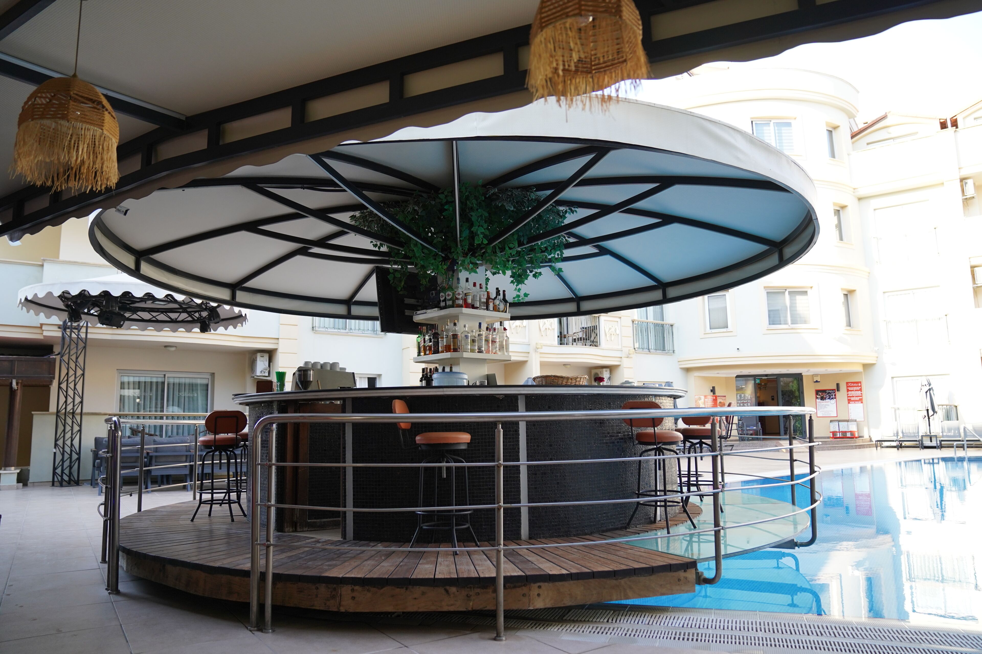 Uma área de bar à beira da piscina com um palco redondo de madeira, banquetes e uma planta pendente.