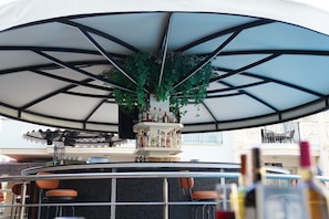 Uma área de bar com um guarda-sol grande, uma planta pendente e diversos frascos.