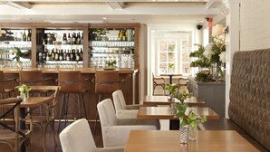 Um restaurante com várias mesas e cadeiras de madeira, um espaço de bar com prateleiras de garrafas e copos, e um banco estofado ao longo da parede.