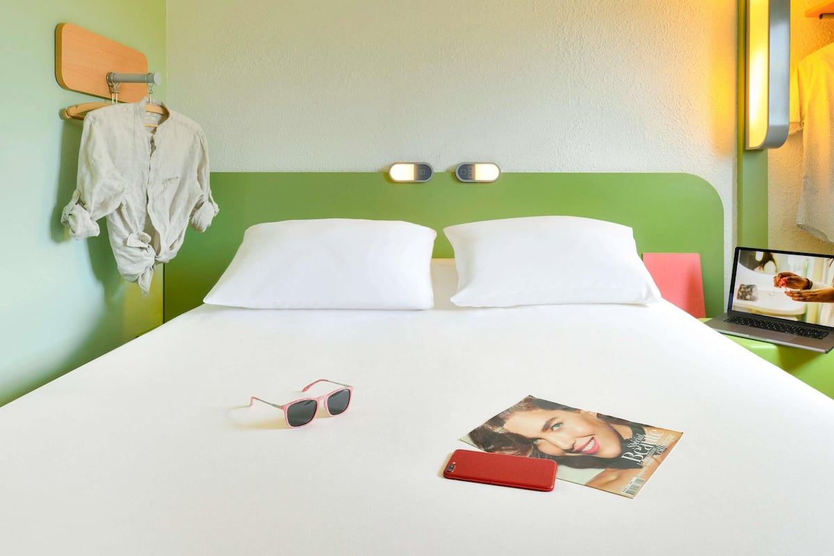 Quarto de hotel com cama, mesa de cabeceira, abajur, porta-toalhas na parede e uma janela.