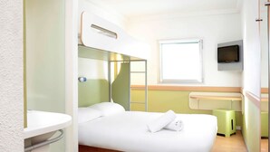 Quarto de hotel compacto com uma cama de solteiro, uma pequena mesa de cabeceira, televisão na parede e uma janela que permite entrada de luz natural.