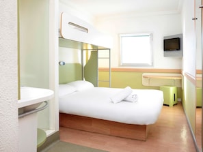 Quarto de hotel compacto com uma cama de solteiro, uma pequena mesa de cabeceira, televisão na parede e uma janela que permite entrada de luz natural.