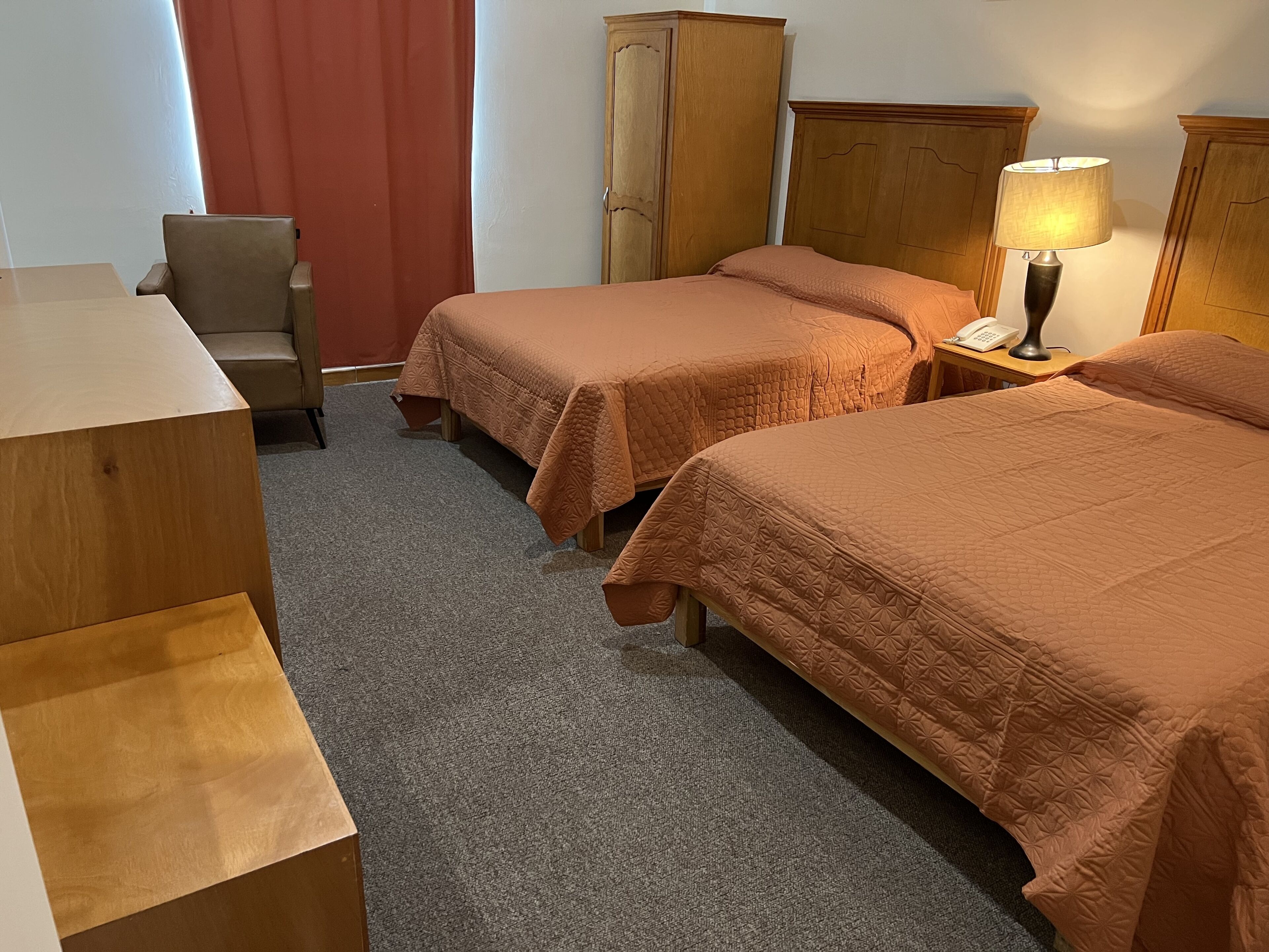 Quarto de hotel com duas camas, uma escrivaninha, uma cadeira, um guarda-roupa e uma mesa de cabeceira com luminária.