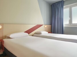 Quarto de hotel com duas camas, um design geométrico na parede, uma janela com cortinas e uma luminária na parede.