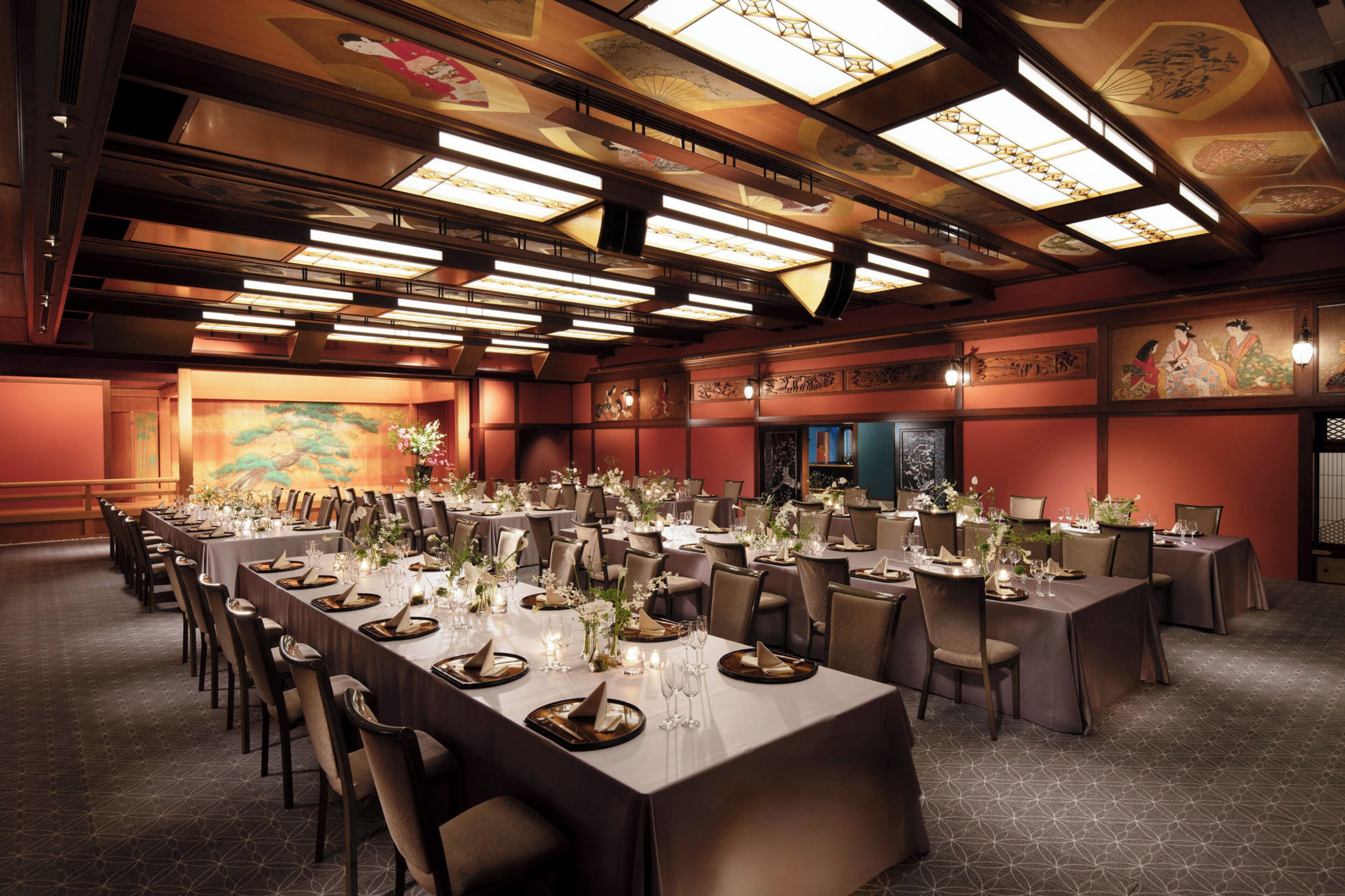 Uma sala de banquetes com longas mesas preparadas para refeições, com decoração tradicional e obras de arte nas paredes.