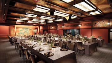 Uma sala de banquetes com longas mesas preparadas para refeições, com decoração tradicional e obras de arte nas paredes.