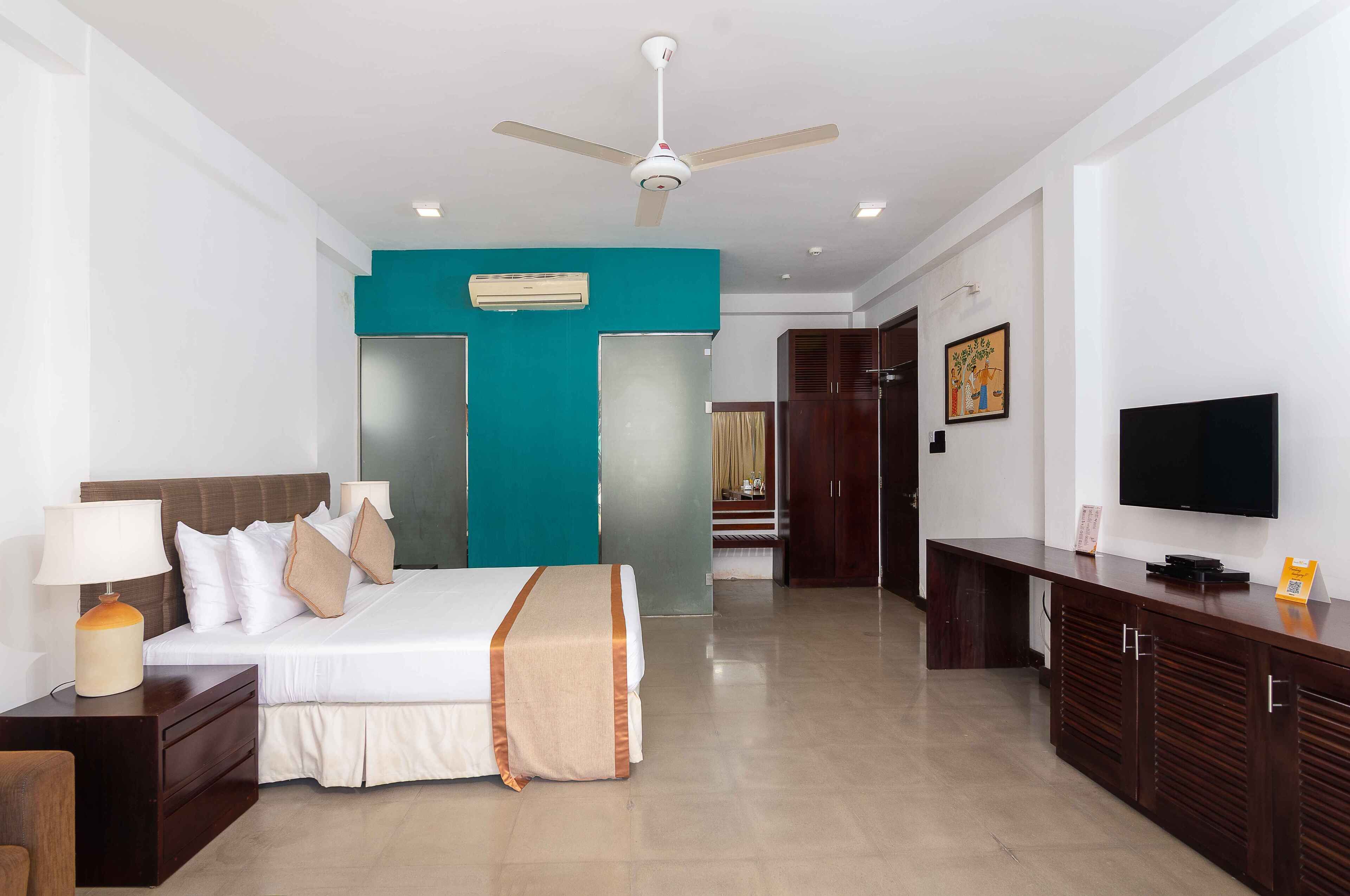 Quarto de hotel com cama, mesa de cabeceira, luminária, ventilador de teto, televisão e escrivaninha com cadeira.