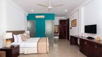 Quarto de hotel com cama, mesa de cabeceira, luminária, ventilador de teto, televisão e escrivaninha com cadeira.