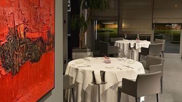 Restaurante com mesas redondas preparadas para refeições, uma grande pintura abstrata na parede e vista para o verde lá fora.