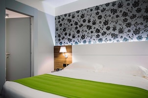 Quarto de hotel com cama, mesa de cabeceira com abajur, papel de parede estampado com flores e uma porta.