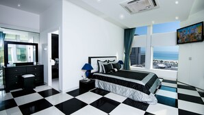 Quarto moderno com piso xadrez, uma cama grande com colcha preta e branca, mesa de cabeceira com abajur e vista da cidade através de amplas janelas.