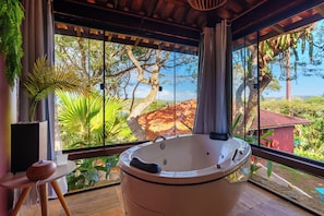 Um banheiro com uma banheira grande, uma mesa pequena com um alto-falante e vista para uma paisagem tropical através de grandes janelas.