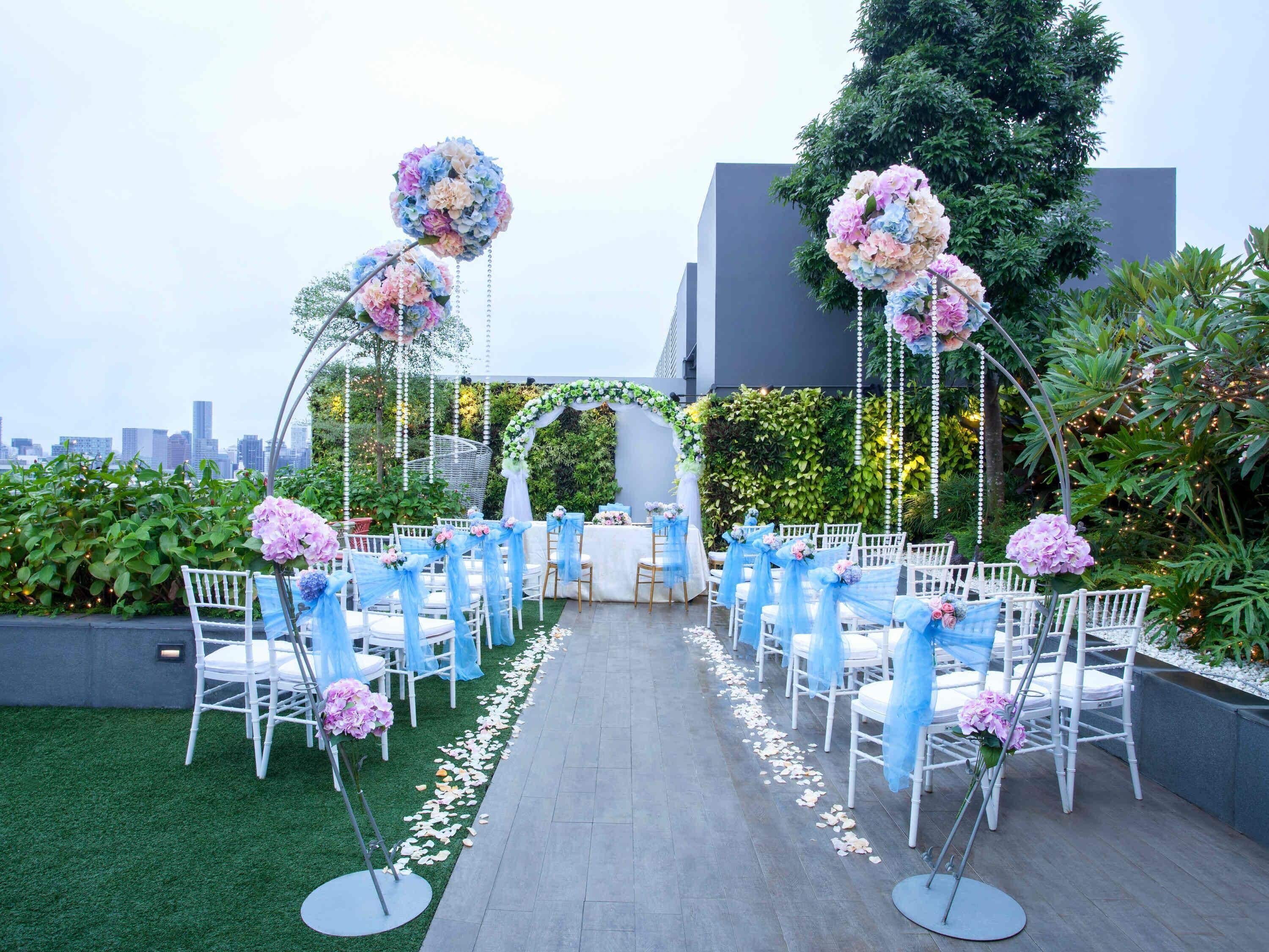 Um espaço para eventos ao ar livre com cadeiras brancas dispostas em fileiras, um arco de flores e vegetação.