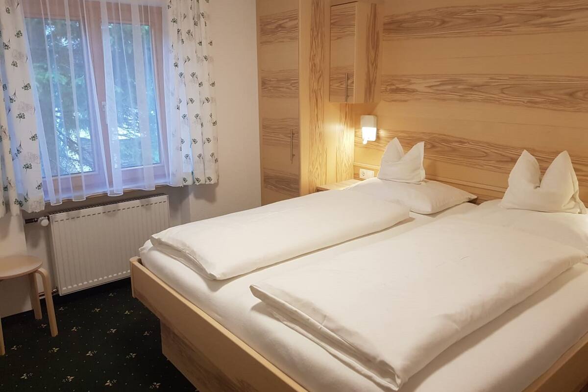 Quarto de hotel com duas camas, uma mesinha, um radiador e uma janela com cortinas finas.