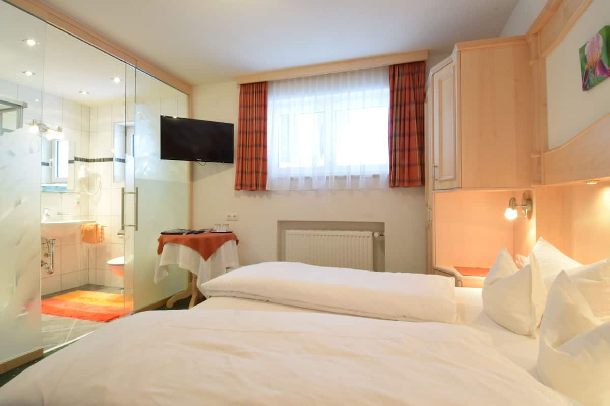 Quarto de hotel com box de vidro, cama com roupa de cama branca, televisão de tela plana na parede e janela com cortinas xadrez.