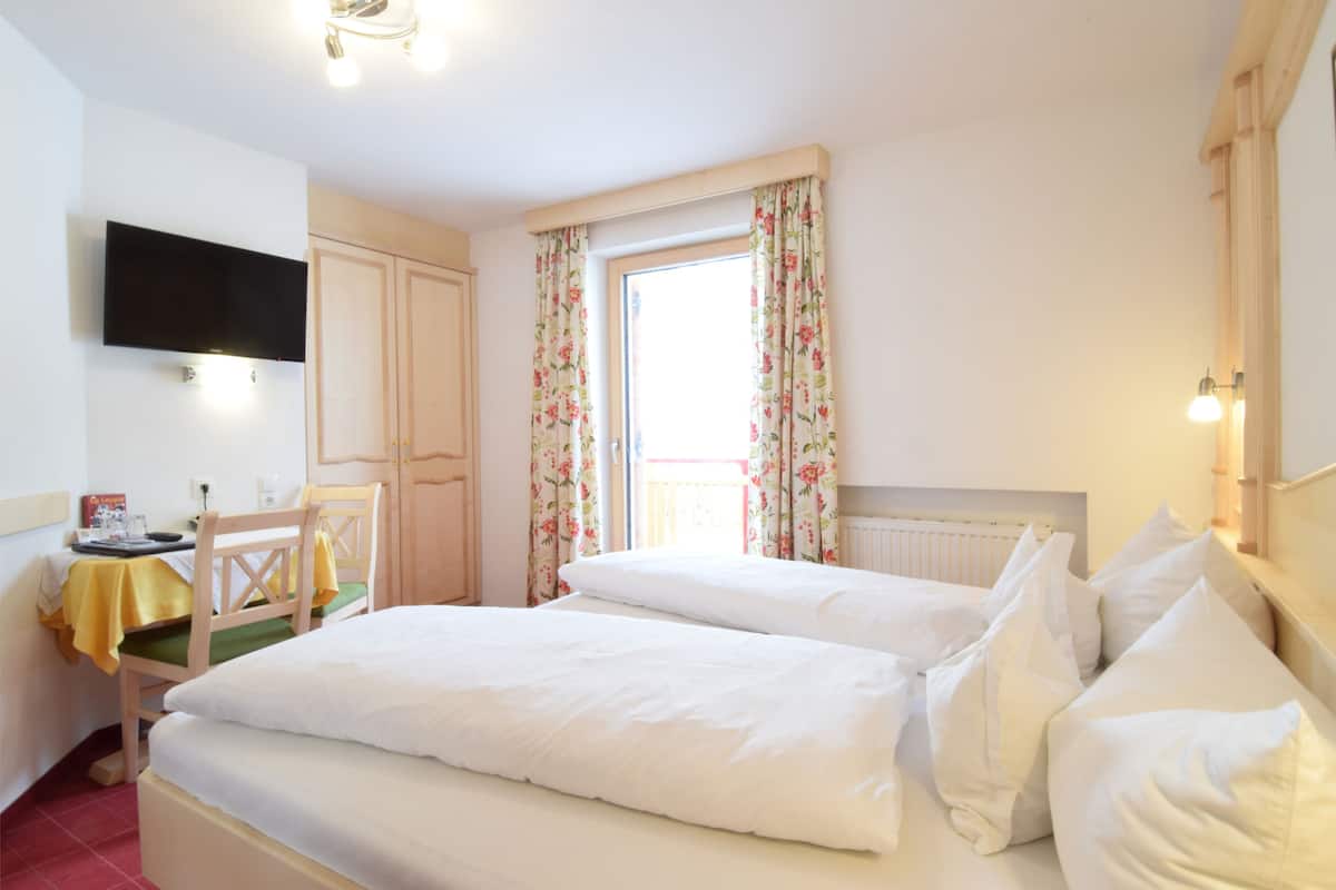 Quarto de hotel com duas camas, uma TV fixada na parede, uma pequena mesa com toalha de mesa amarela e uma janela com cortinas florais.
