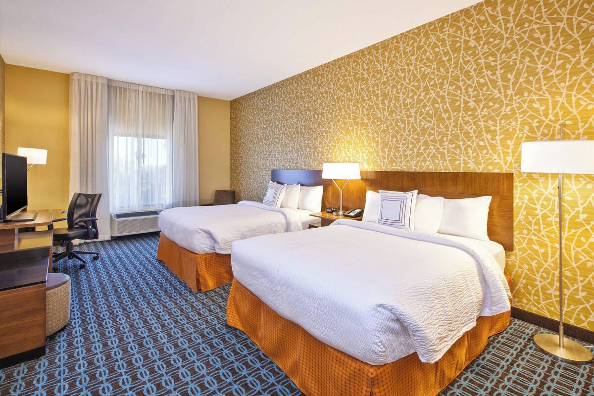 Quarto de hotel com duas camas, uma escrivaninha com cadeira, televisão e carpete estampado.