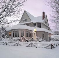 Uma casa grande de dois andares com telhado inclinado, rodeada por árvores cobertas de neve e uma cerca.